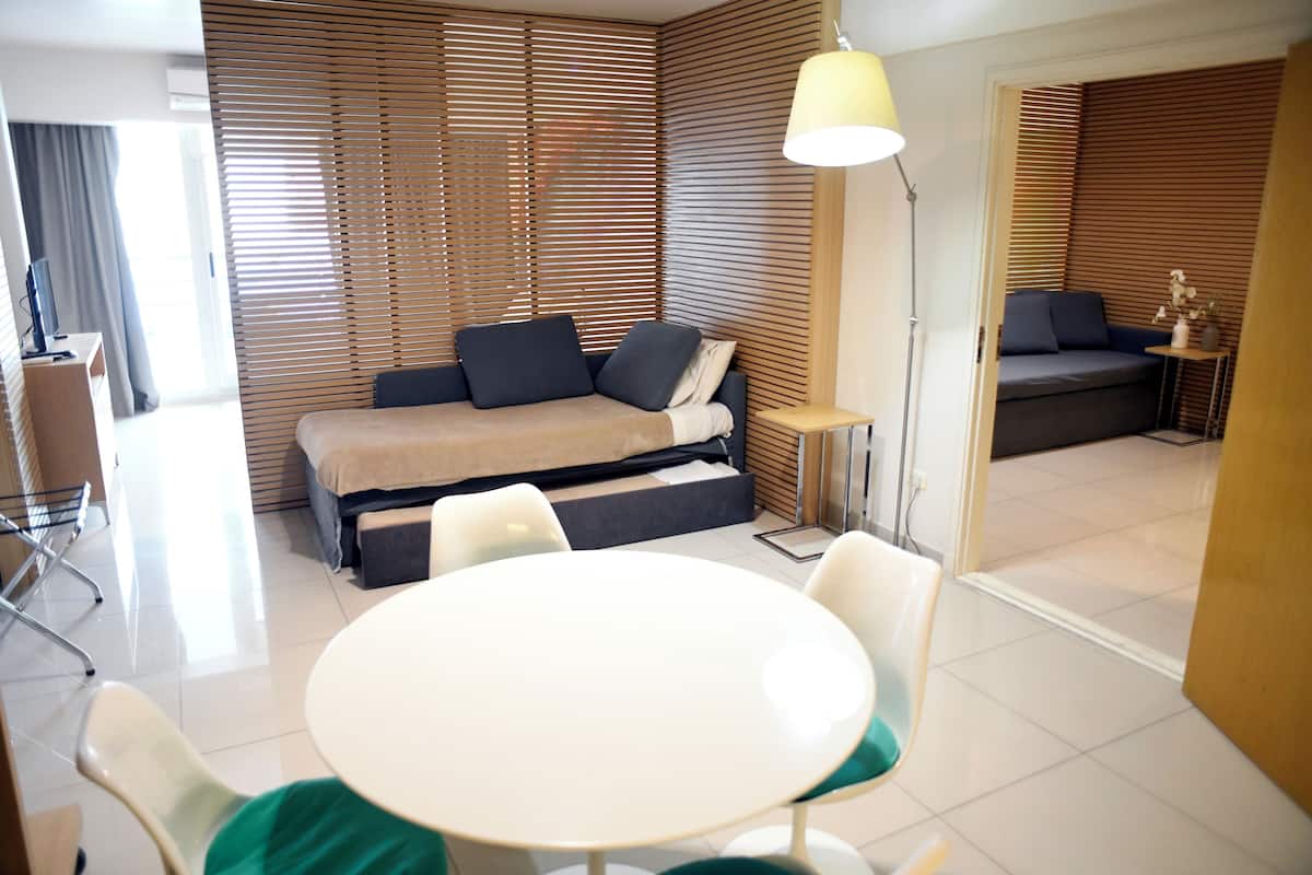 Quarto de hotel moderno com sofá-cama, mesa de jantar com cadeiras e pequena mesa lateral com abajur.