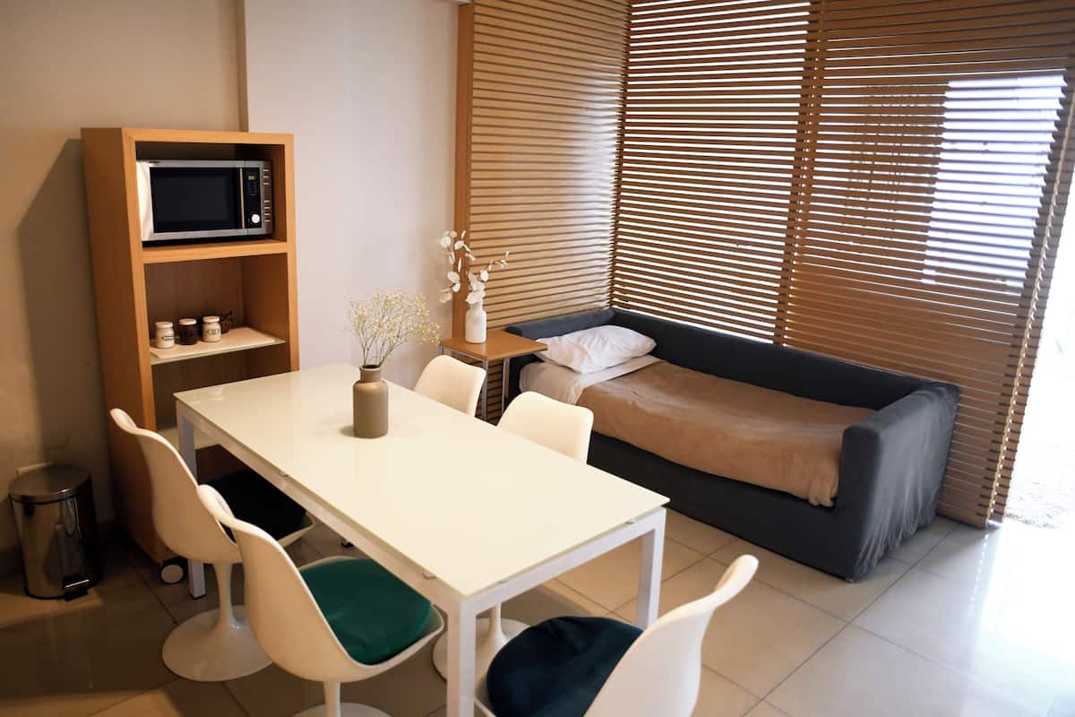 Uma sala de estar moderna com uma mesa de jantar branca, cadeiras brancas, um sofá cinza e uma estante de madeira com um micro-ondas.