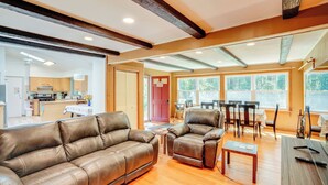 Uma sala de estar ampla com sofás de couro, um espaço de jantar com cadeiras de madeira, uma cozinha ao fundo e grandes janelas que proporcionam luz natural.