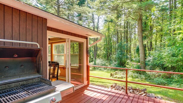 Um deck de madeira com churrasqueira, uma porta de vidro que dá para um pátio e vista para uma área arborizada.