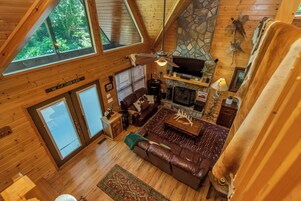 Interior de uma cabana com lareira de pedra, sofá de couro e uma janela ampla com vista para vegetação.