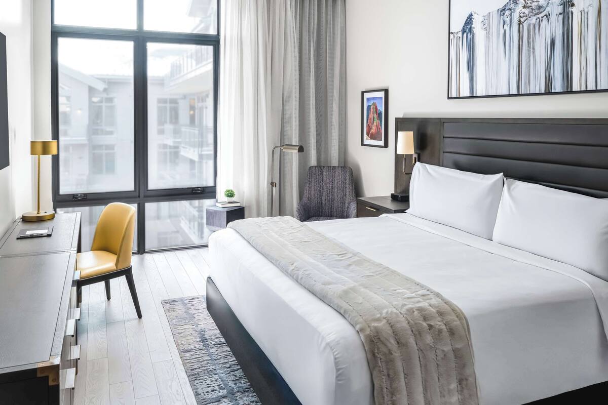 Quarto de hotel moderno com uma cama grande, uma escrivaninha, uma cadeira e vista para edifícios pela janela.