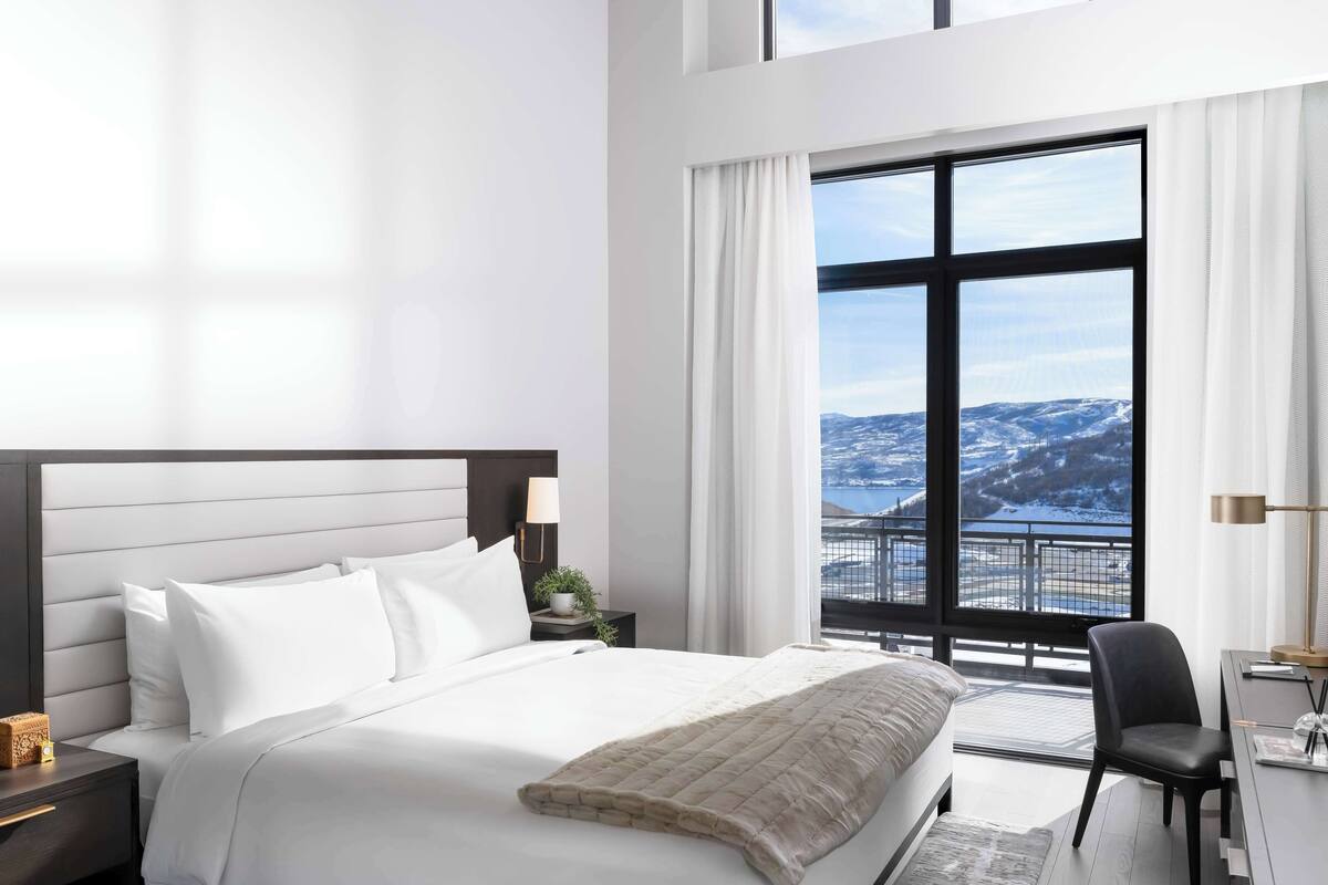 Quarto de hotel moderno com uma cama grande, uma mesa de escritório e vista para uma paisagem nevada através de uma grande janela.