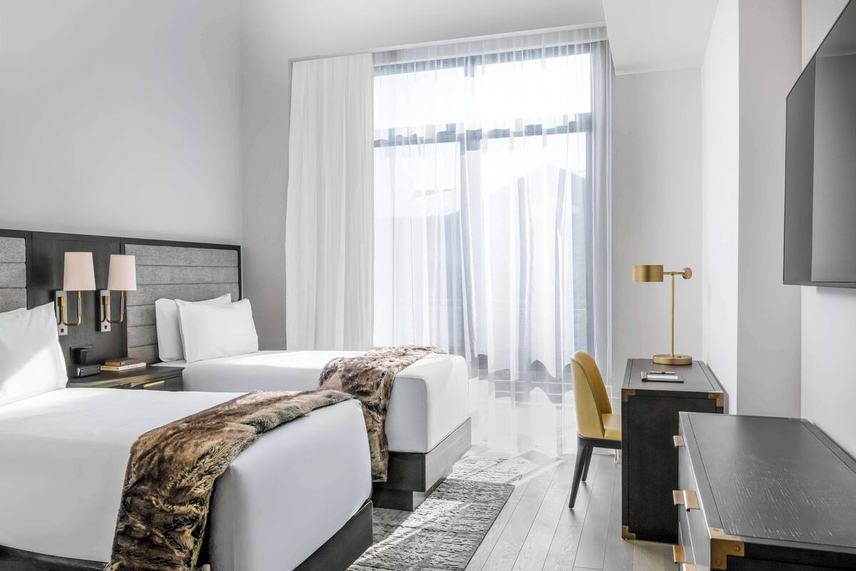 Quarto de hotel moderno com duas camas, uma escrivaninha, uma cadeira e uma janela grande com cortinas leves.