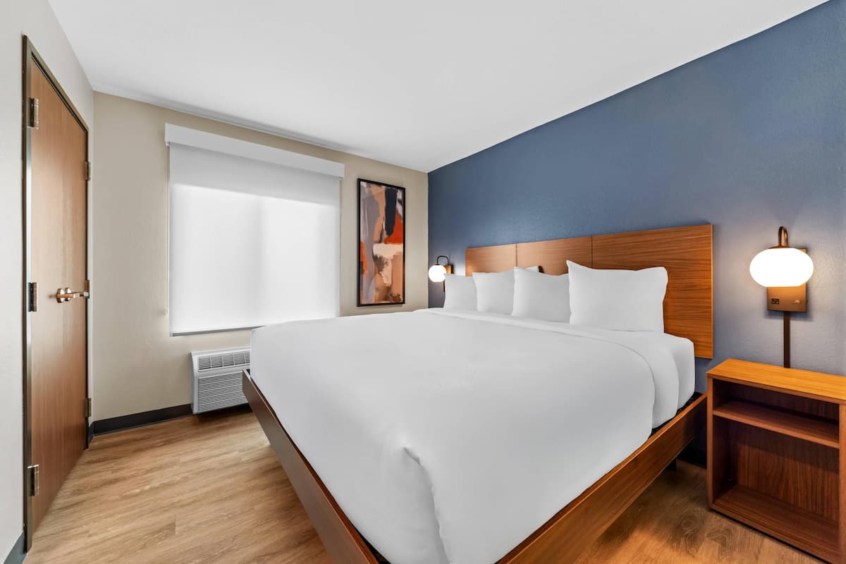 Quarto de hotel com uma cama grande, uma mesinha de cabeceira, uma luminária na parede e uma janela com persianas.
