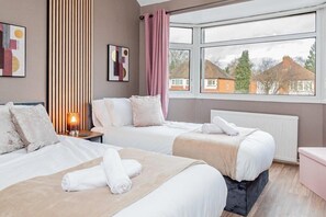 Quarto de hotel com duas camas, uma janela grande com vista para casas, uma mesa de cabeceira com abajur e uma cortina rosa.