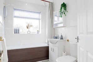 Casa de banho moderna com banheira, sanita e lavatório. Tem uma janela com cortinas decorativas e uma planta em vaso.