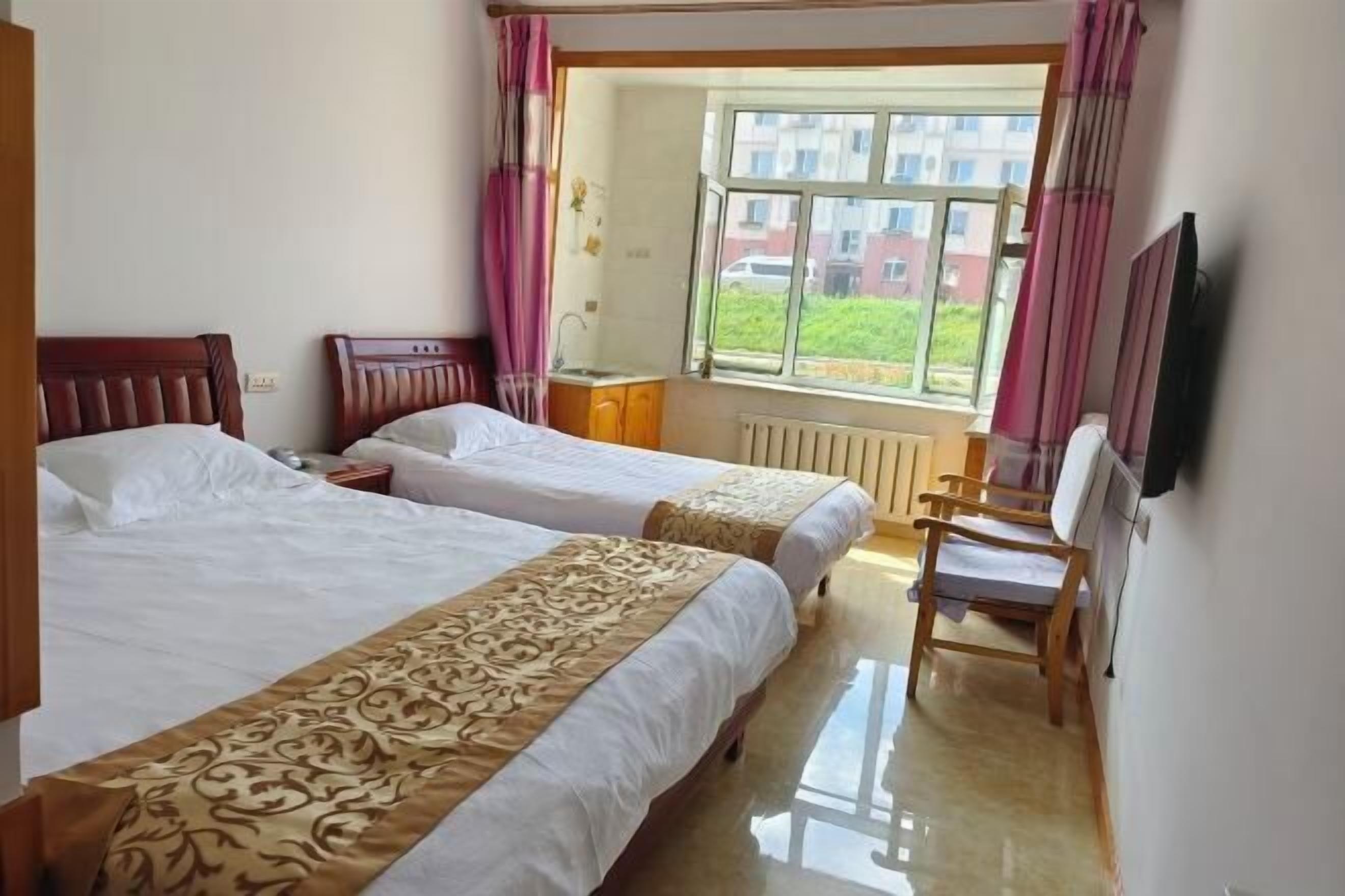 Quarto de hotel com duas camas, janela com cortinas, uma cadeira de madeira, radiador e televisão na parede.