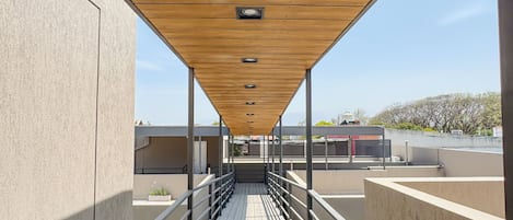 Uma galeria coberta ao ar livre, com teto de madeira, grades de metal e vista para os arredores.