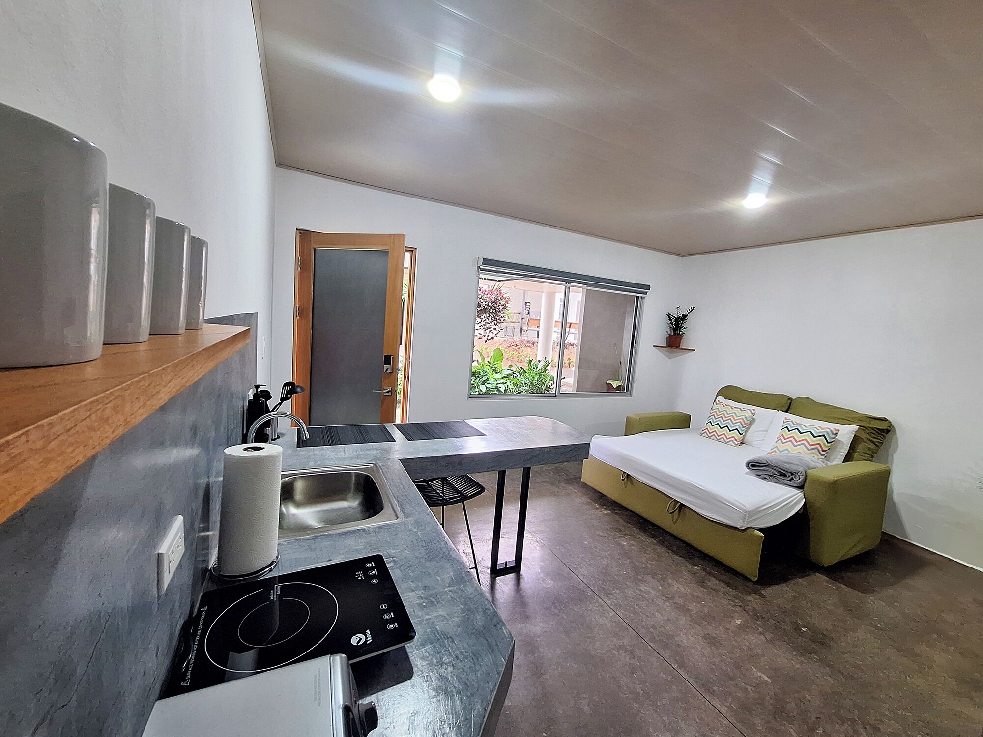 Cozinha compacta com pia, fogão e bancada. Há uma janela com vista para o verde lá fora. Um sofá e uma pequena mesa estão na área de estar adjacente.