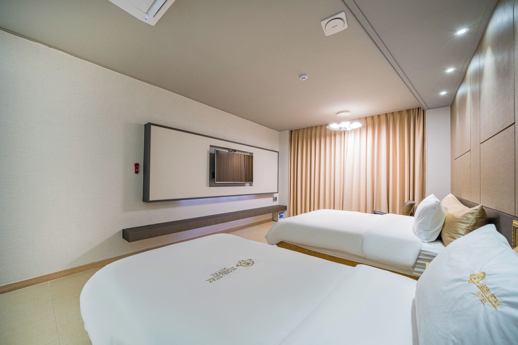 Quarto de hotel com duas camas, uma televisão montada e uma janela grande com cortinas.