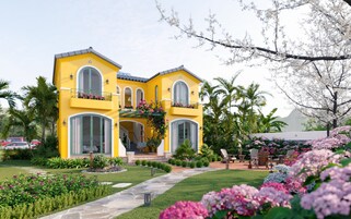 Uma casa amarela de dois andares com varanda, rodeada por vegetação exuberante e plantas floridas.
