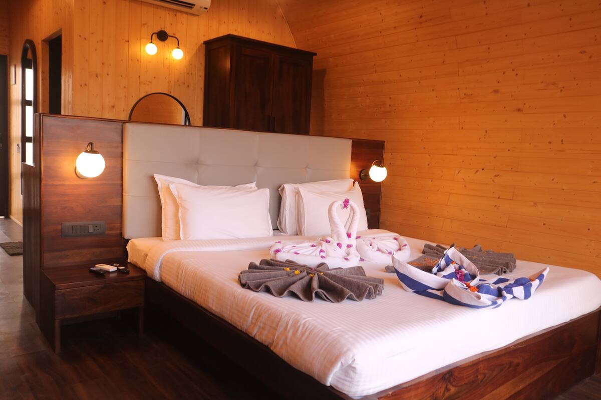 Quarto de hotel com decoração em madeira, uma cama grande com roupa de cama branca, uma mesa de cabeceira com abajur e um guarda-roupa de madeira.