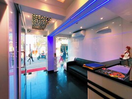 Uma área de recepção moderna com entrada por porta de vidro, um sofá de couro preto e um painel de menu digital.