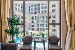 Quarto com uma grande janela com vista para um pátio que inclui piscina e edifícios. Há duas cadeiras e uma mesa com uma planta e um menu.