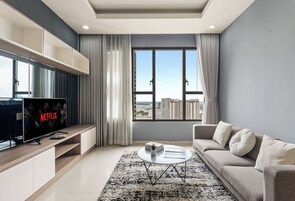 Uma sala de estar moderna com uma televisão grande, um sofá, uma mesa de centro e vista para a cidade através de amplas janelas.