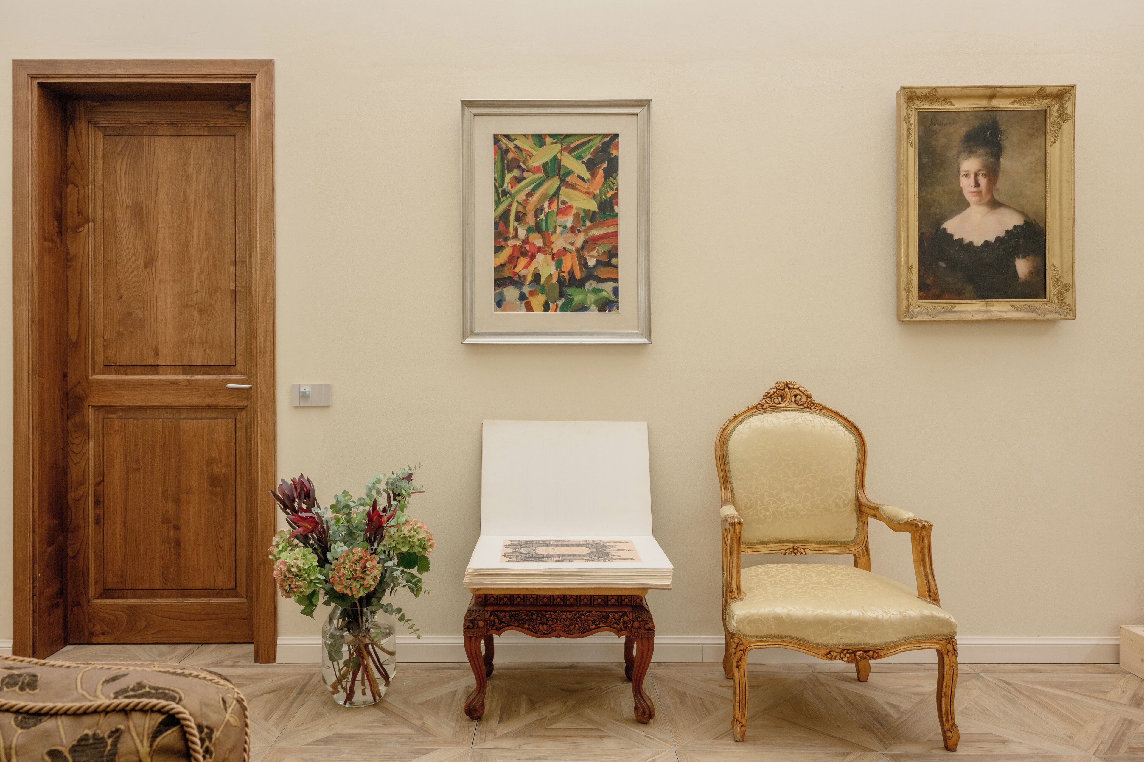 Um quarto com porta de madeira, uma pintura abstrata emoldurada, um retrato emoldurado, um cavalete branco, uma cadeira decorativa e um vaso com flores.