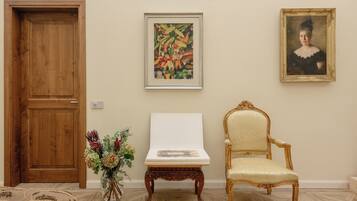 Um quarto com porta de madeira, uma pintura abstrata emoldurada, um retrato emoldurado, um cavalete branco, uma cadeira decorativa e um vaso com flores.
