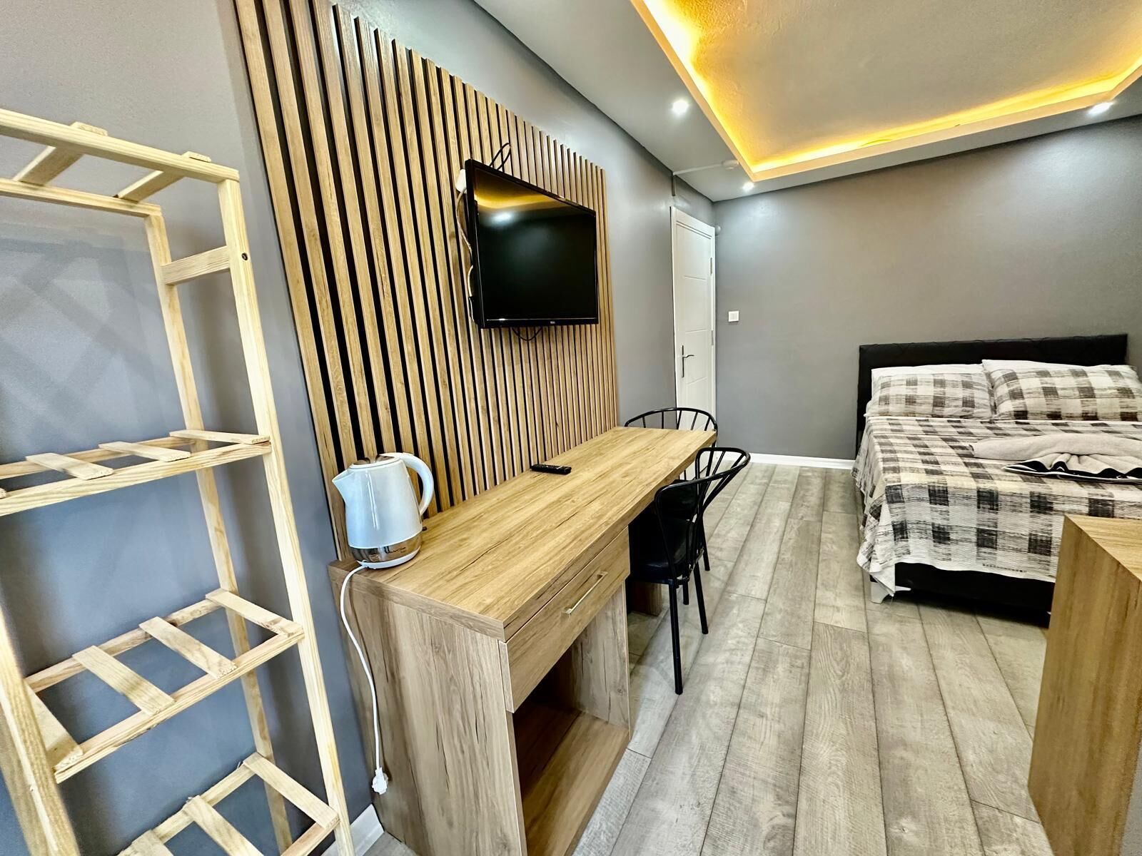 Um quarto moderno com escrivaninha de madeira, televisão fixada na parede, cama com colcha xadrez e chão de madeira.