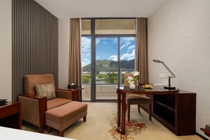 Quarto de hotel com uma janela grande, uma mesa com luminária, uma cadeira e uma cama.