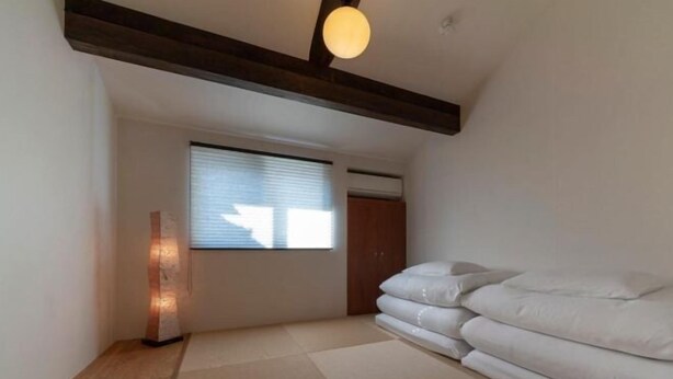 Quarto minimalista com tatame, uma janela grande com persianas, um abajur e roupa de cama organizada.