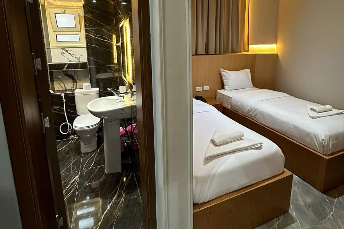 Quarto de hotel compacto com banheiro, duas camas de solteiro, penteadeira com espelho e área de chuveiro.