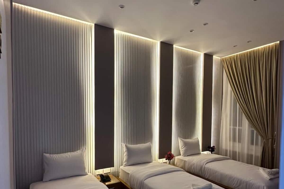 Quarto de hotel com três camas de solteiro, cabeceira de madeira, mesa de cabeceira e uma janela grande com cortinas.