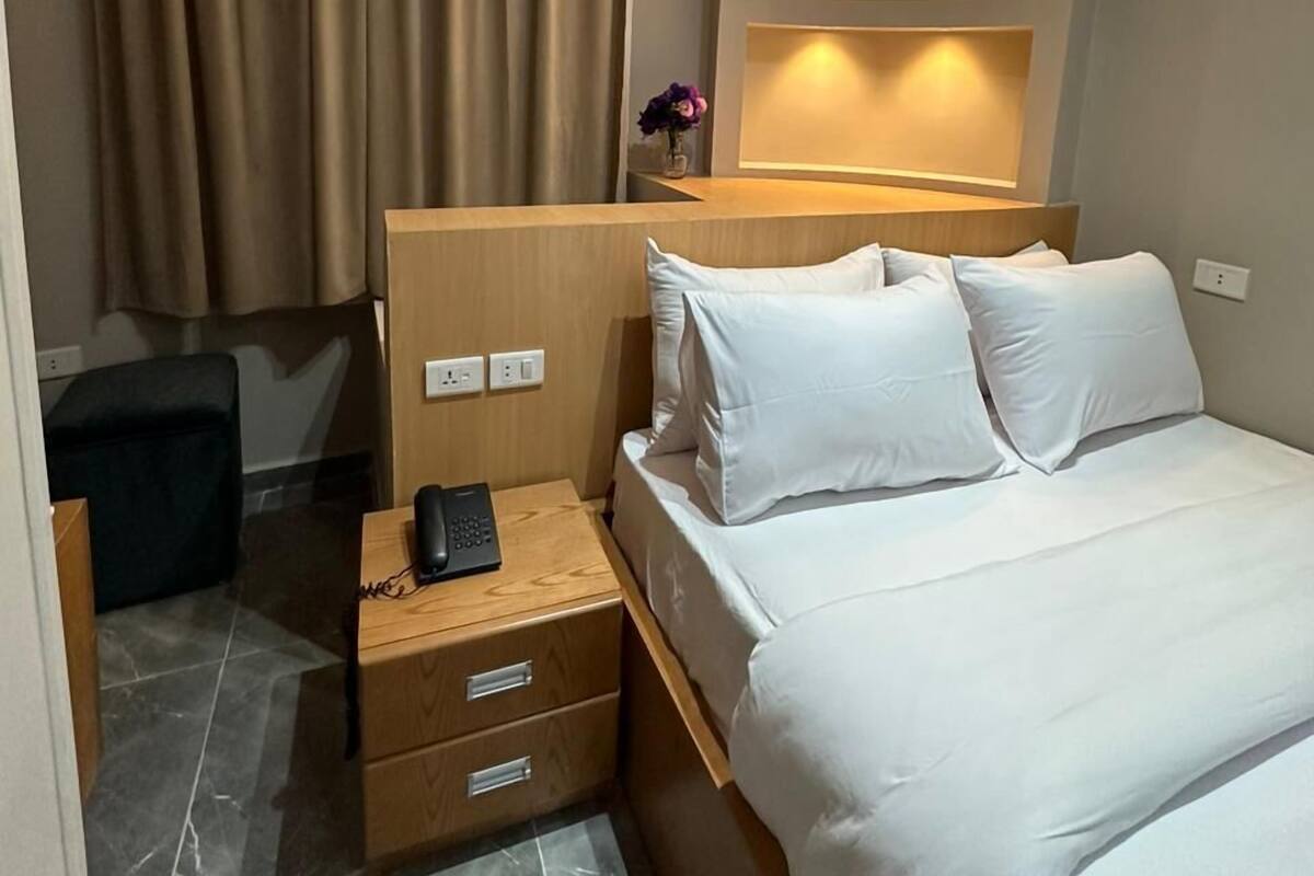 Quarto de hotel com cama, mesa de cabeceira, luminária e prateleira na parede com um vaso.