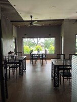 Uma área de refeições com mesas e cadeiras, janelas amplas com vista para o verde e um ventilador de teto.