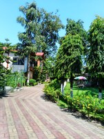 Uma trilha pavimentada leva a um edifício com telhado vermelho, cercado por vegetação exuberante e um gramado bem cuidado.