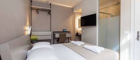 Quarto de hotel moderno com cama, escrivaninha, poltrona, televisão e chuveiro.