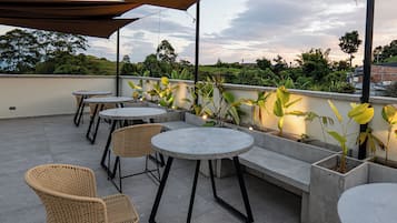 Terraço no telhado com área de estar externa, incluindo mesas redondas e cadeiras de vime, cercado por plantas em vasos e vista para a paisagem.