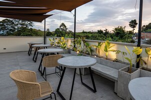 Terraço no telhado com área de estar externa, incluindo mesas redondas e cadeiras de vime, cercado por plantas em vasos e vista para a paisagem.