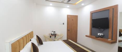 Quarto de hotel com cama, ventilador de teto, televisão fixada na parede, uma mesinha com cadeira e porta.