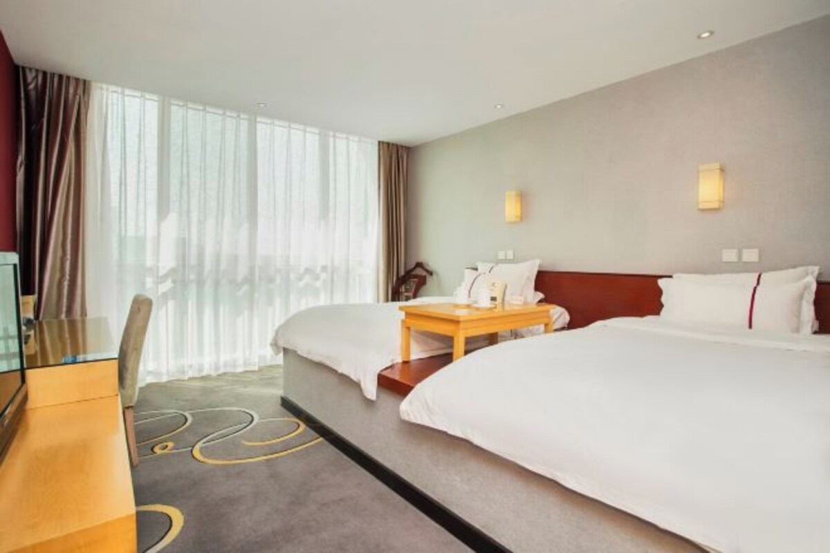 Quarto de hotel com duas camas, uma escrivaninha, uma cadeira, uma mesa e uma janela grande com cortinas.