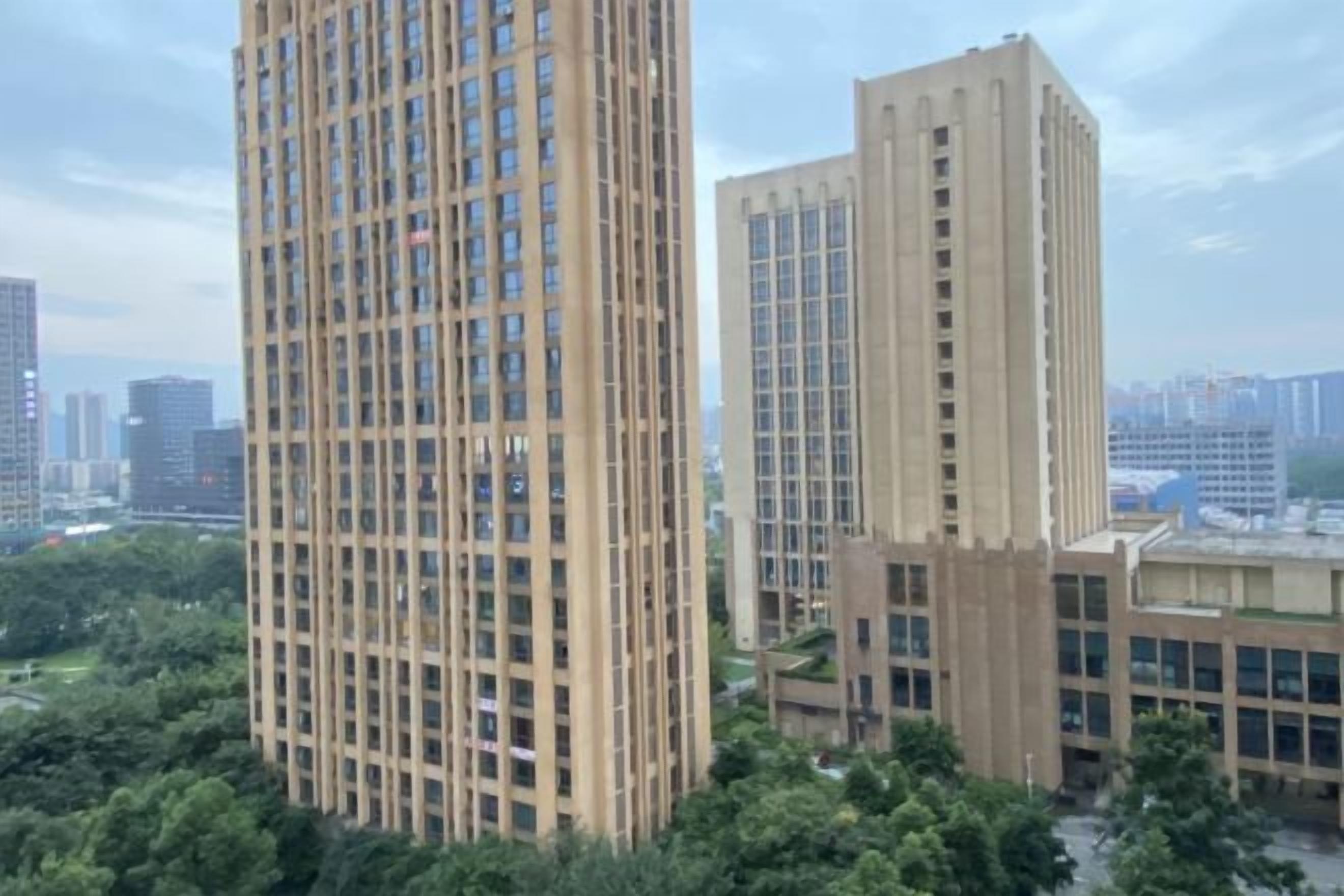 Um edifício alto com fachada em forma de grade, cercado por vegetação e outros edifícios.