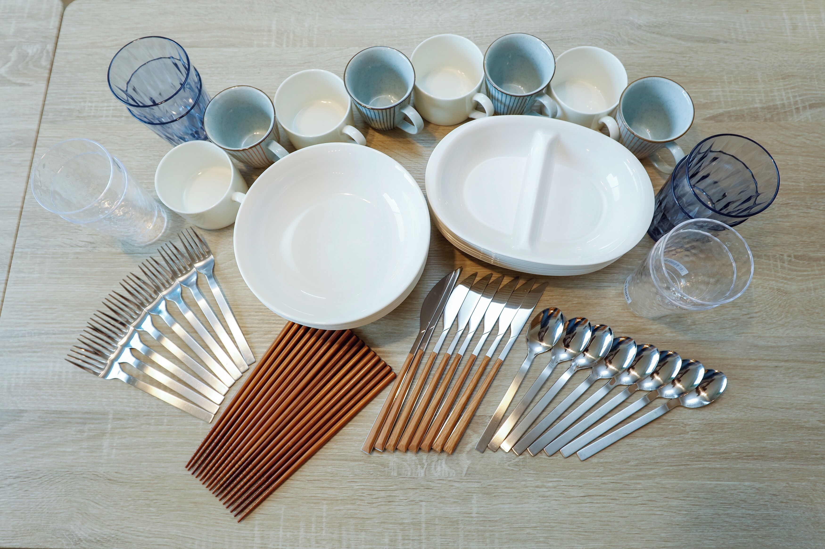 Mesa posta com pratos brancos, tigelas, talheres e copos de vidro azuis.
