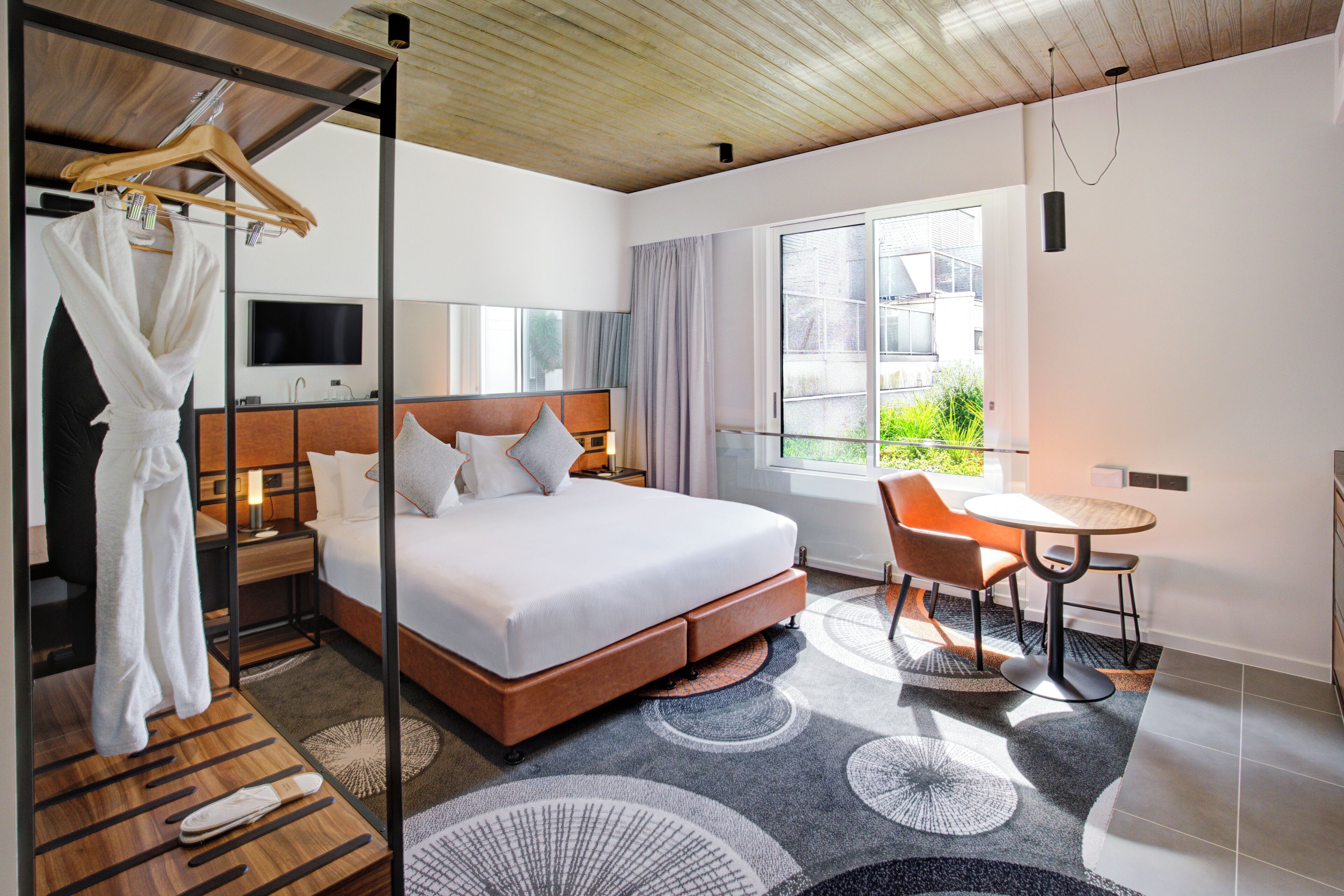 Quarto de hotel moderno com uma cama grande, uma escrivaninha com cadeira, televisão e vista para o verde lá fora.