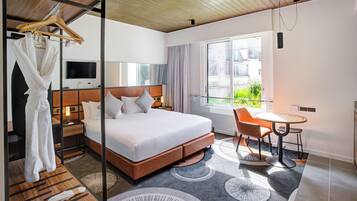 Quarto de hotel moderno com uma cama grande, uma escrivaninha com cadeira, televisão e vista para o verde lá fora.
