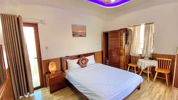 Quarto de hotel com cama, cabeceira de madeira, mesa de cabeceira com luminária, pequena mesa com cadeiras e janela com cortinas.