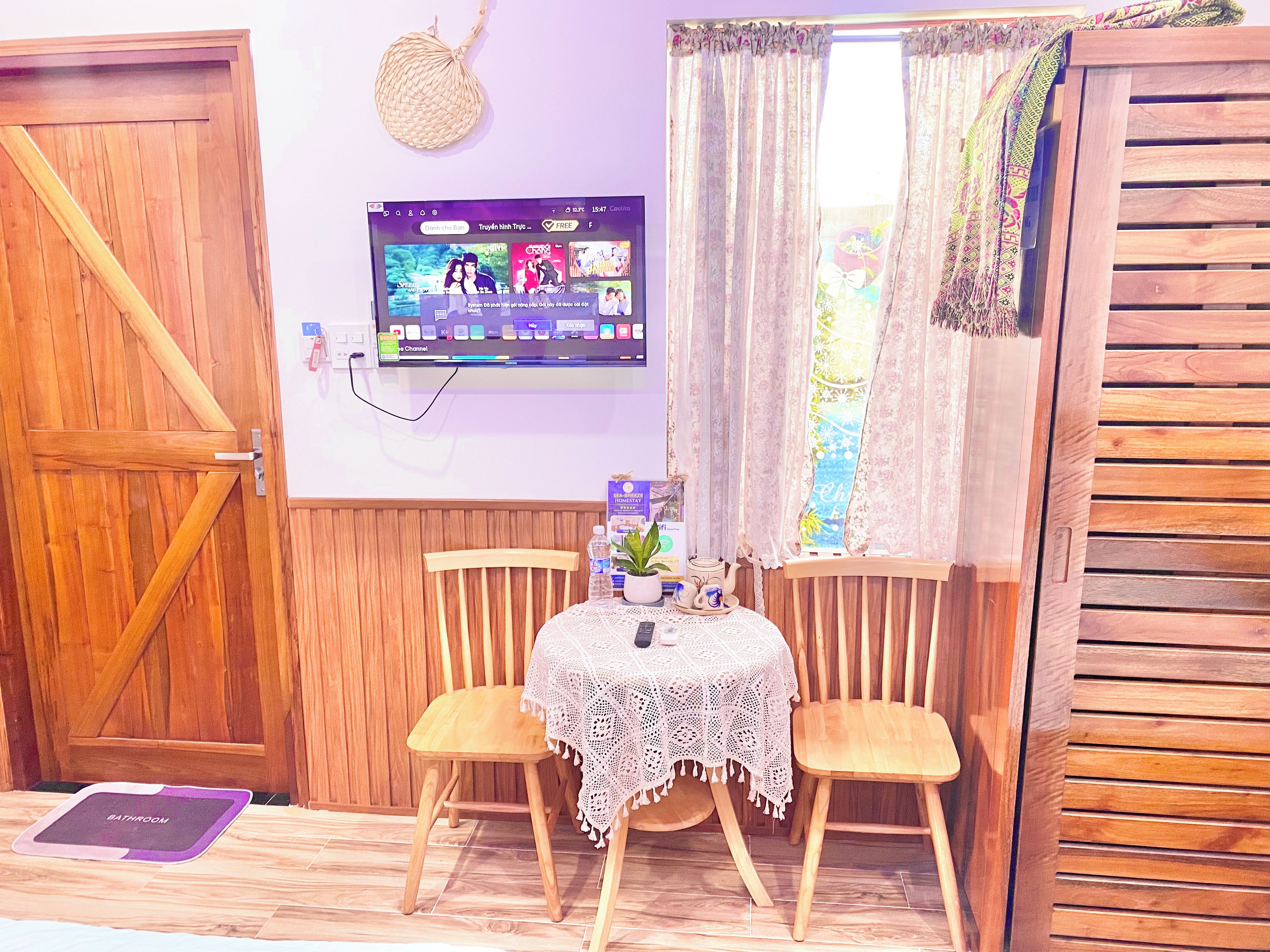 Quarto com porta de madeira, televisão fixada na parede, mesa pequena com toalha de renda, duas cadeiras de madeira e janela com cortinas.