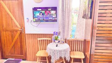 Quarto com porta de madeira, televisão fixada na parede, mesa pequena com toalha de renda, duas cadeiras de madeira e janela com cortinas.