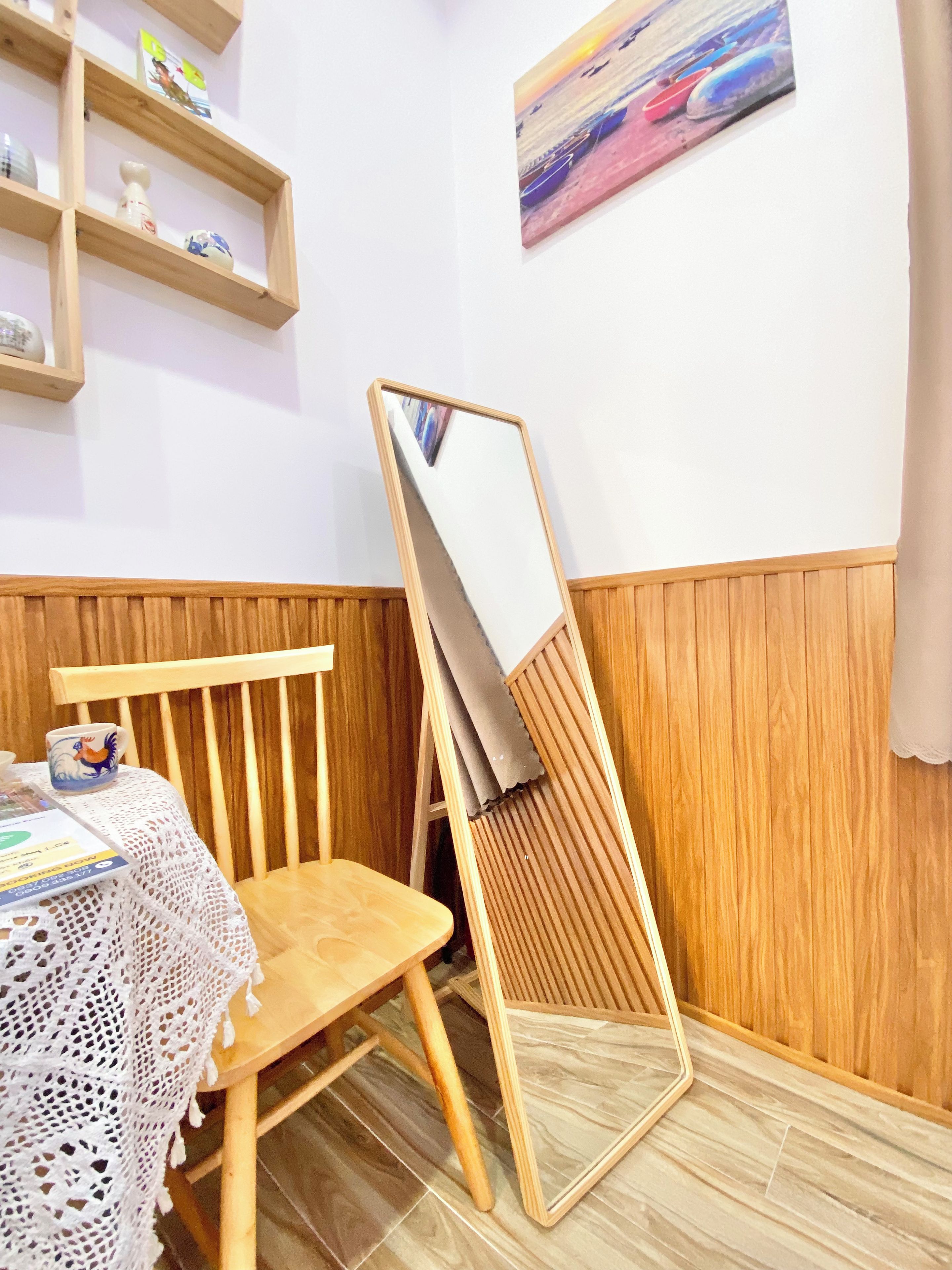 Um quarto com uma cadeira de madeira, uma mesa com toalha de renda branca, uma estante com objetos decorativos e um quadro na parede.