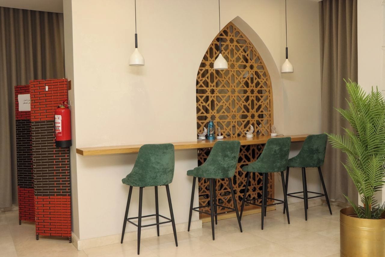 Uma área de bar com balcão de madeira, banquetas verdes, luminárias pendentes e um extintor fixado num armário vermelho.