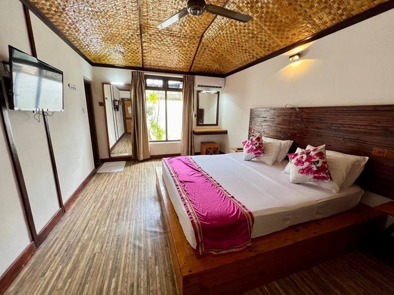 Um quarto com cama de madeira, ventilador de teto, televisão e janela com cortinas.