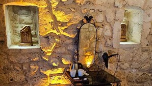 Quarto com paredes de pedra, lareira, suporte de metal para chávenas e duas janelas pequenas decoradas.