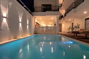 Uma piscina com deck de madeira e espreguiçadeiras, uma varanda com guarda-corpo e uma área de cozinha visível através de uma divisória.