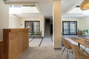 Um interior espaçoso com uma mesa de recepção de madeira, uma área de jantar com cadeiras de vime e uma escada que dá acesso a um andar superior.