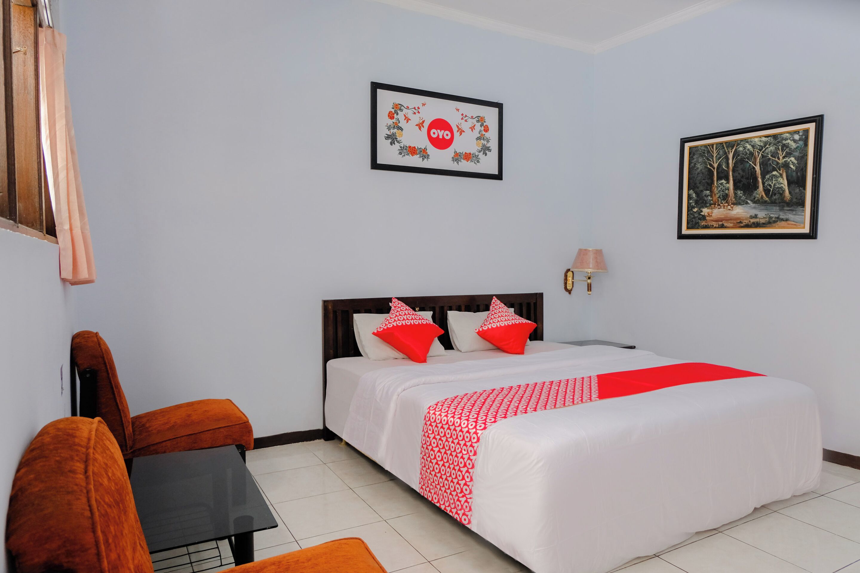 Quarto de hotel com uma cama, uma escrivaninha e uma cadeira. Há um quadro na parede e uma luminária na escrivaninha.