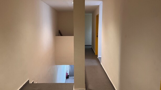 Um corredor estreito e bem iluminado, com um pilar central, que leva a um quarto com uma porta no final.